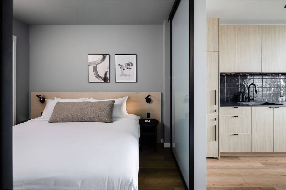 Quarto de hotel moderno com uma cama, mesa de cabeceira e vista para a área da cozinha adjacente.