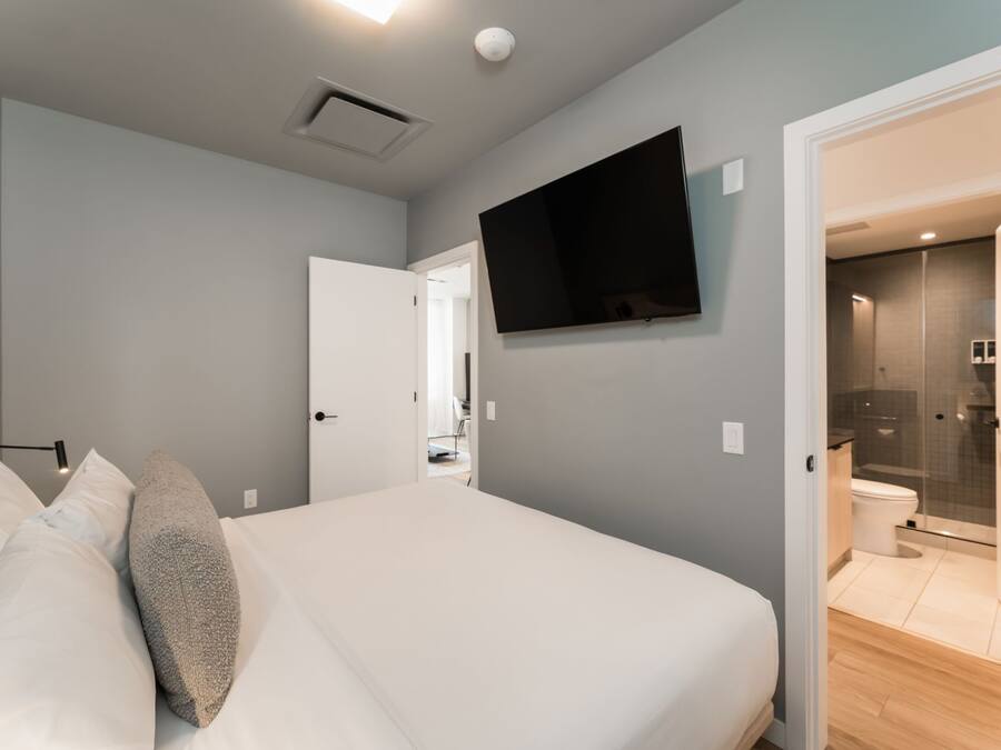 Quarto moderno com cama, TV de tela plana na parede e banheiro visível com box de vidro.