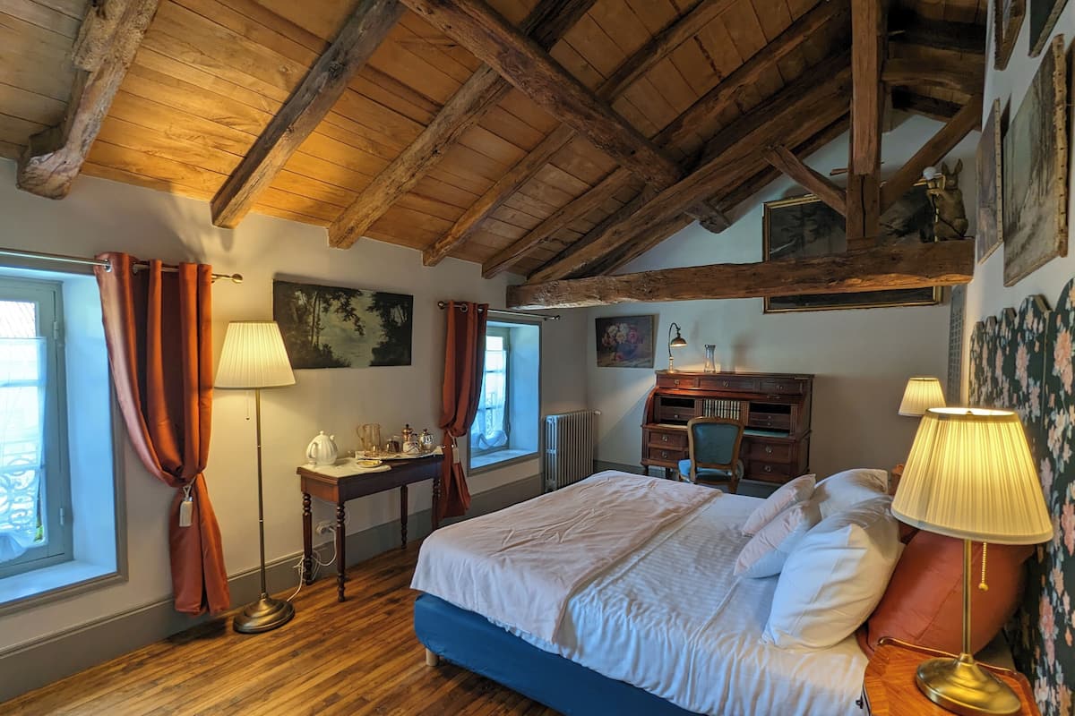 Quarto com teto de madeira, cama com roupa branca, cómoda de madeira e janela com cortinas.
