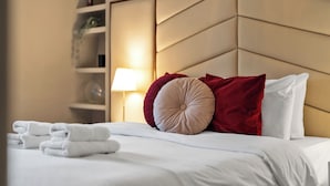 Cama bem arrumada com lençóis brancos, um travesseiro vermelho e um travesseiro bege. Toalhas brancas dobradas estão sobre a cama.