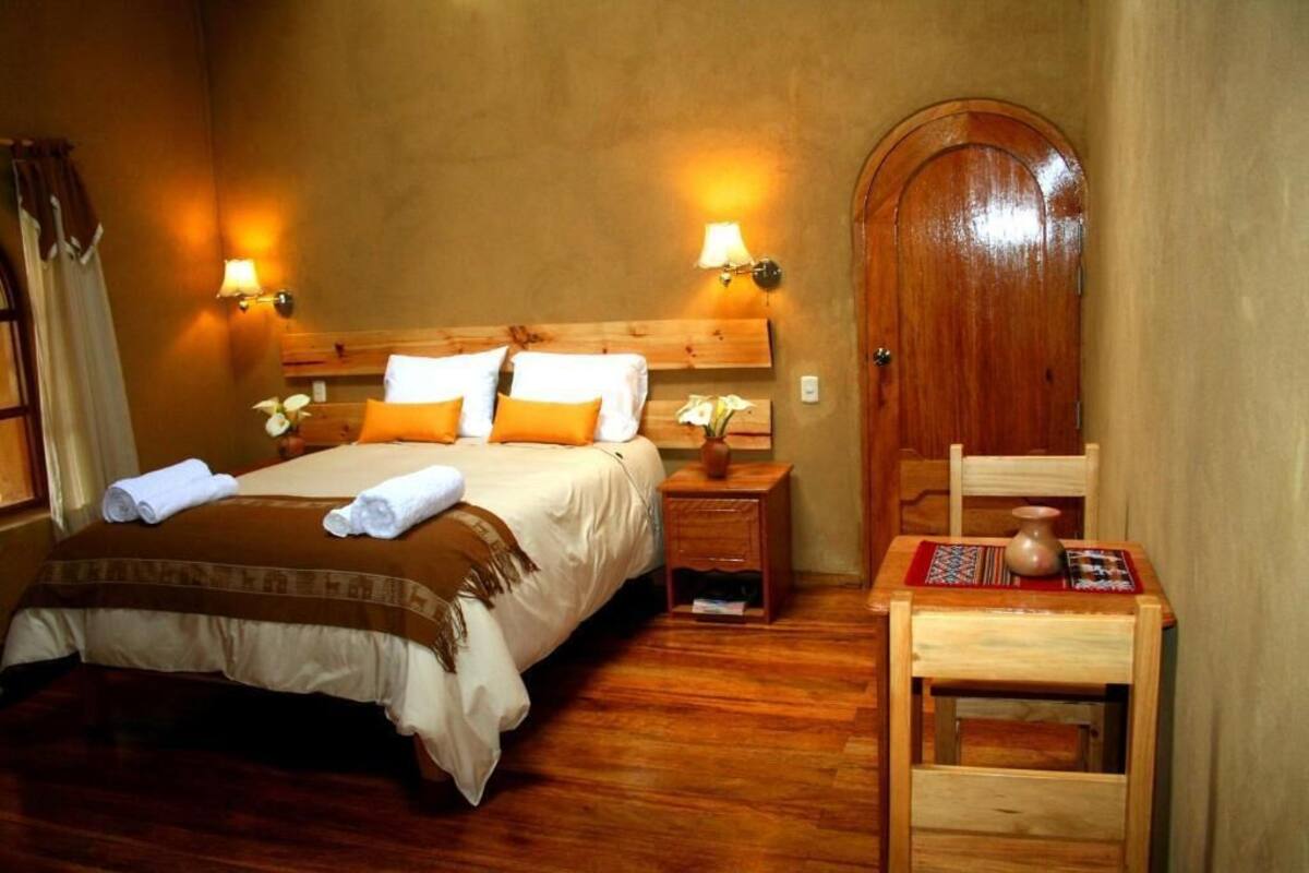 Quarto de hotel com cama de madeira, duas luminárias de cabeceira, mesa de cabeceira de madeira, uma pequena mesa com um vaso e uma porta de madeira.