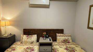 Quarto de hotel com duas camas de solteiro, uma mesa de cabeceira, uma luminária, uma pequena mesa com um vaso de flores e um aparelho de ar condicionado na parede.