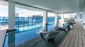 Piscina infinita no terraço com espreguiçadeiras, deck de madeira e vista para o horizonte da cidade.