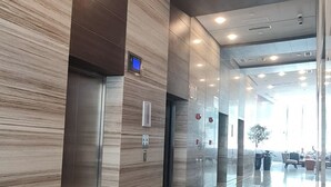 Uma área de elevador moderna com piso polido, painel digital e porta de vidro que dá para um espaço interior iluminado.