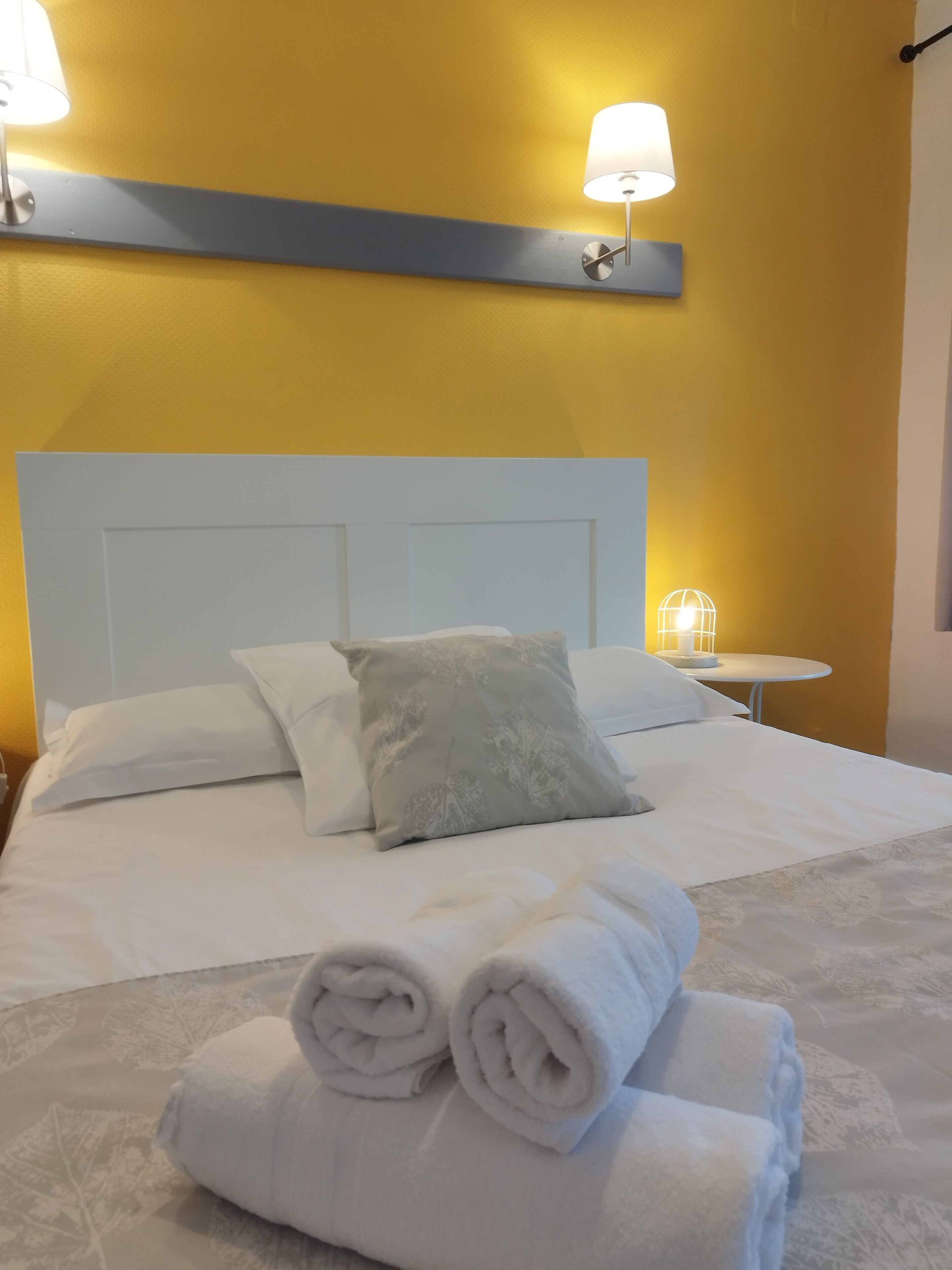 Cama bem arrumada com lençóis brancos e toalhas dobradas, cabeceira branca e duas luminárias fixadas na parede.