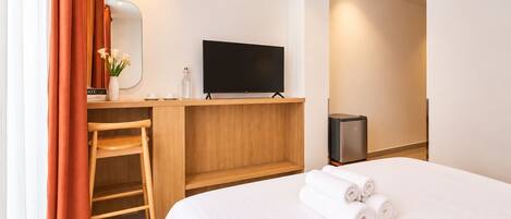 Quarto de hotel com cama, escrivaninha, televisão e lixeira.