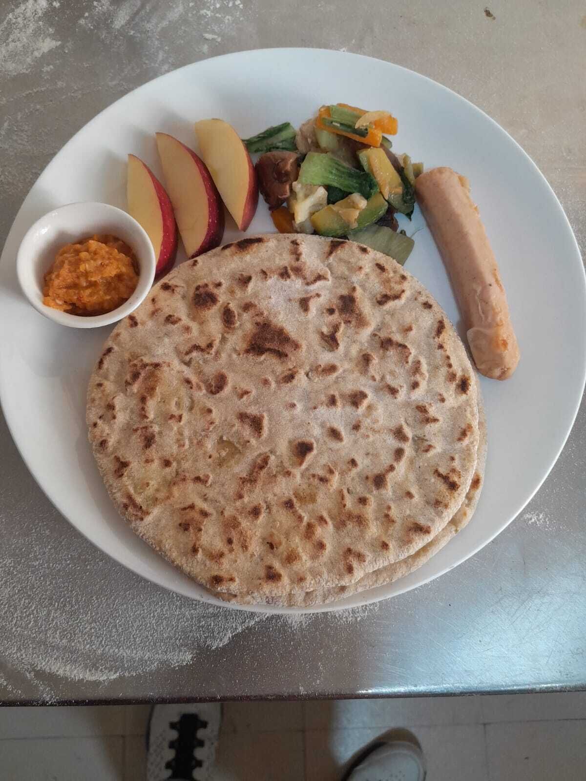 Um prato com pão achatado, fatias de maçã, linguiça, um misto de legumes e um pequeno recipiente com chutney.