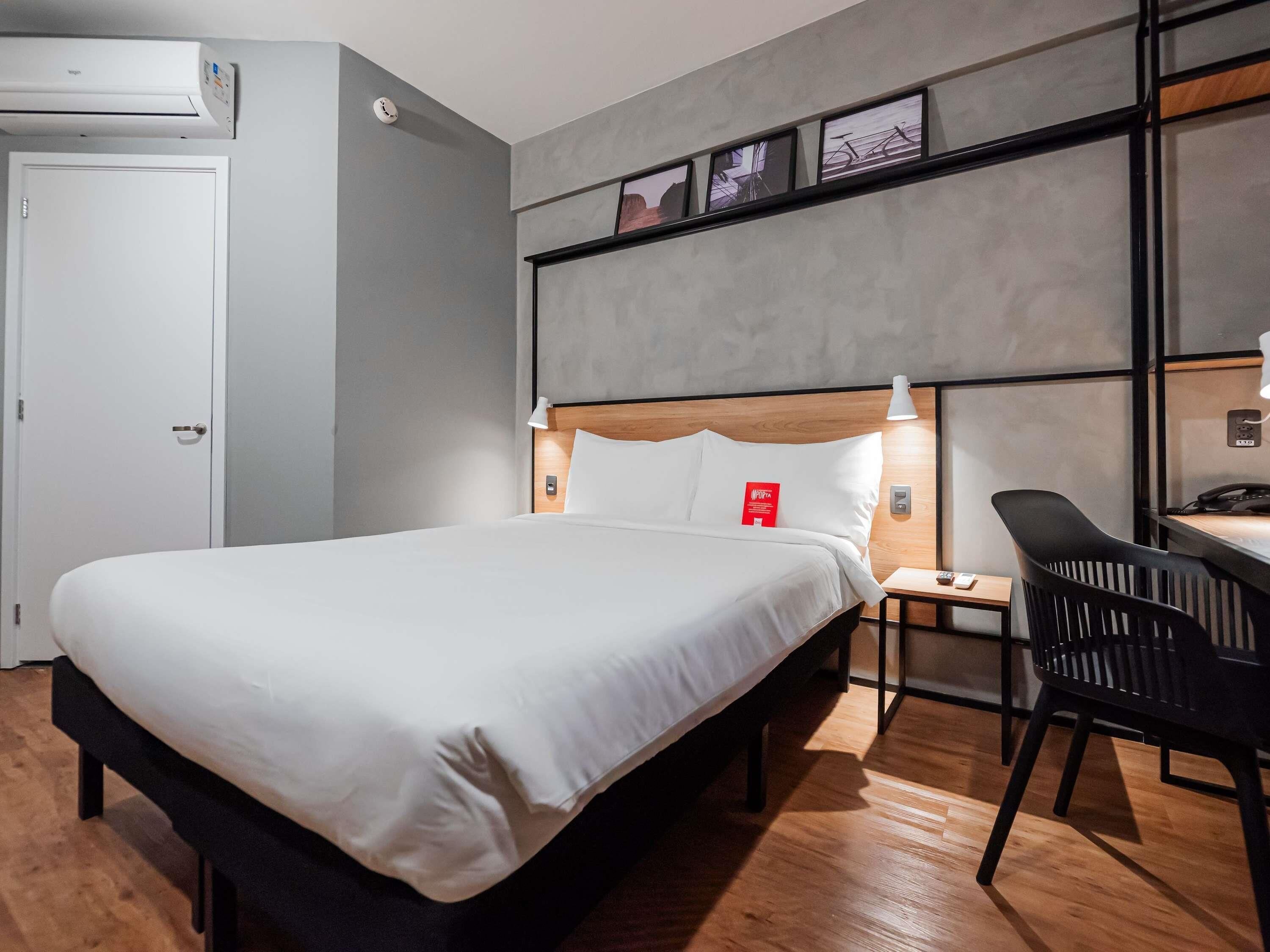 Quarto de hotel com cama, escrivaninha, cadeira, prateleira na parede com fotografias e uma porta.