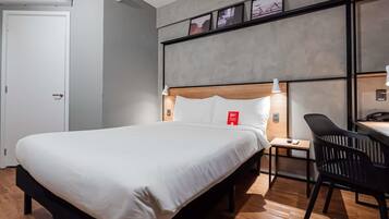 Quarto de hotel com cama, escrivaninha, cadeira, prateleira na parede com fotografias e uma porta.