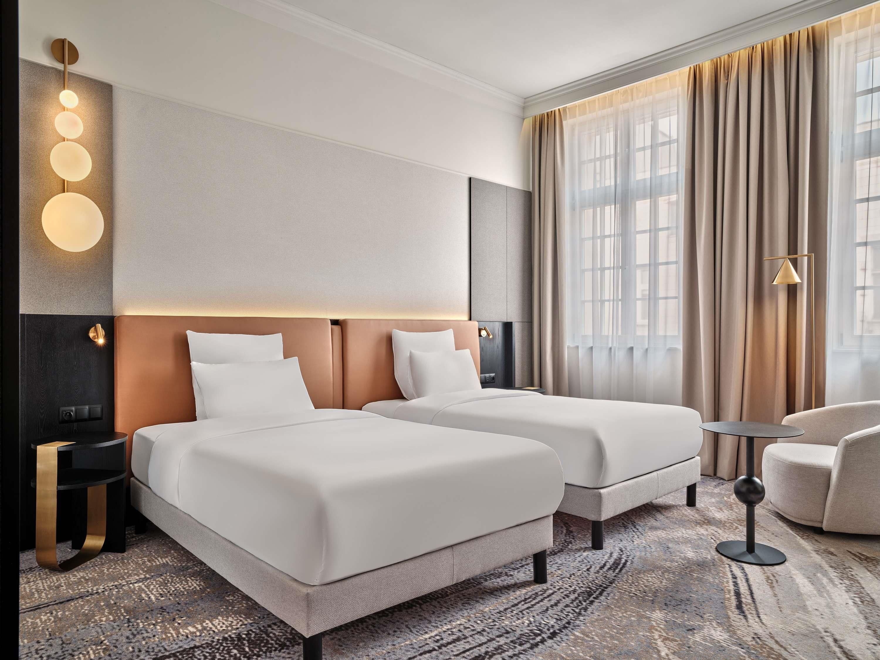 Quarto de hotel com duas camas, uma janela grande com cortinas, uma mesa redonda e uma luminária moderna.