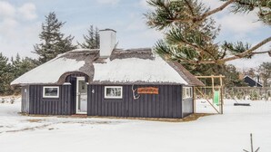 Uma pequena cabana de madeira escura com telhado coberto de neve e chaminé, rodeada por árvores.