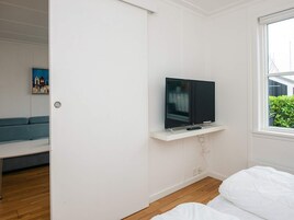 Quarto moderno com televisão na parede branca, piso de madeira e janela com vista para vegetação.