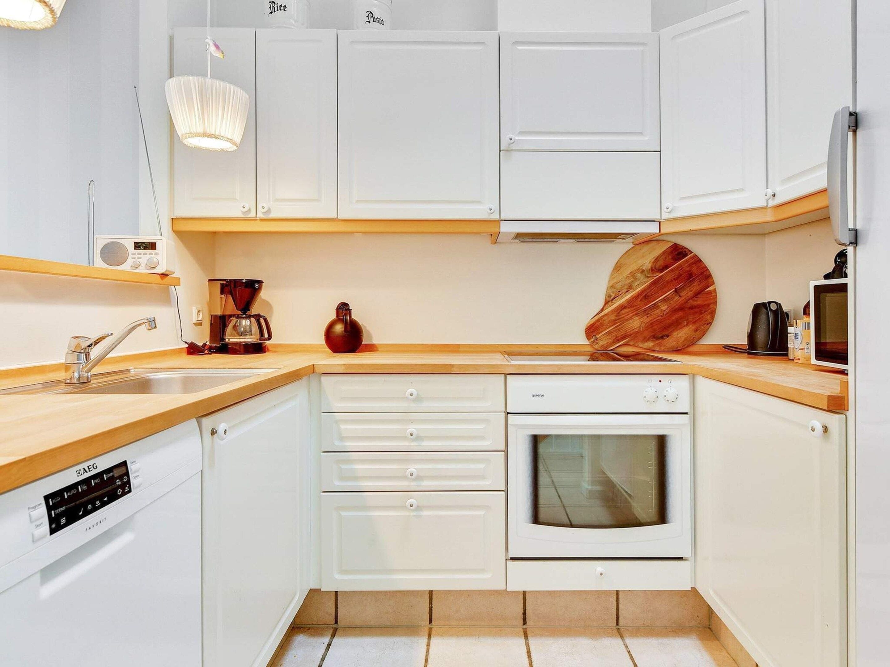 Cozinha compacta com armários brancos, bancada de madeira, forno embutido, cafeteira e rádio.