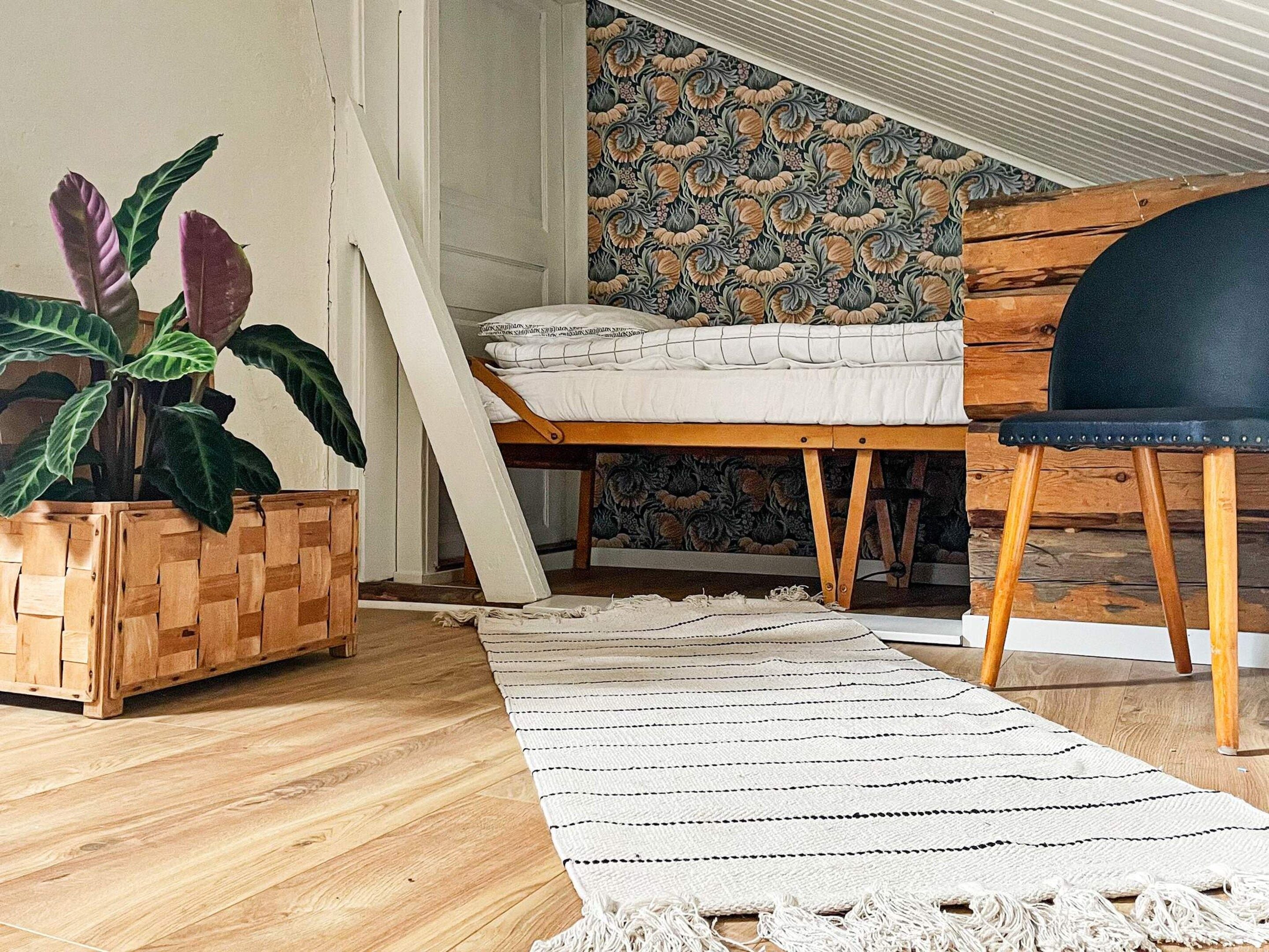 Quarto com piso de madeira, cama com colchão branco, cadeira preta, tapete tecido e planta em vaso.