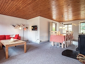 Um quarto acolhedor com teto de madeira, um sofá vermelho, uma mesa de jantar com cadeiras, uma lareira e uma TV fixada na parede.