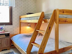 Uma cama de solteiro com escada de madeira, abajur e uma pequena mesa de cabeceira com um vaso de flores.