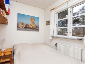 Quarto com cama, janela com cortinas, radiador, quadro na parede e uma pequena mesa de cabeceira.
