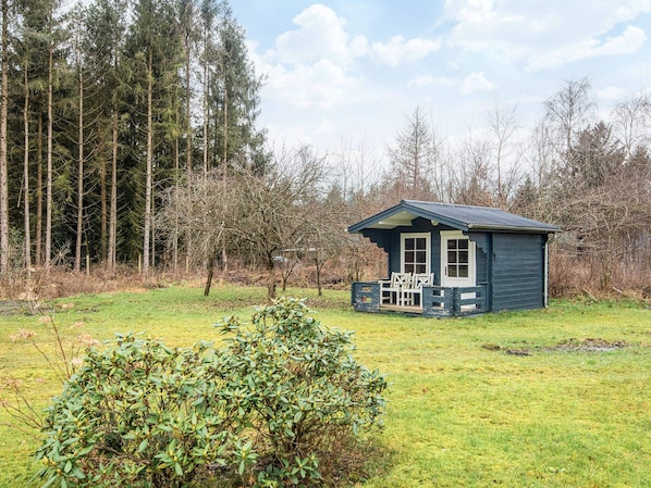 Uma cabana pequena com varanda, cercada por um campo gramado e árvores densas.