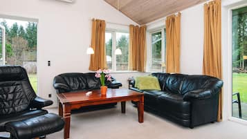 Uma sala de estar moderna com um sofá de couro preto, uma mesa de centro de madeira e uma poltrona de couro preto. Há uma janela com cortinas, um ventilador de teto e um aparelho de ar condicionado.