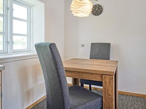 Uma sala de jantar com mesa de madeira e duas cadeiras, janela com moldura branca, relógio na parede e luminária.