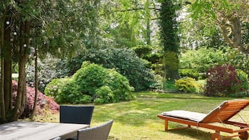 Um jardim com mesa e cadeiras, espreguiçadeira e diversas árvores e arbustos.