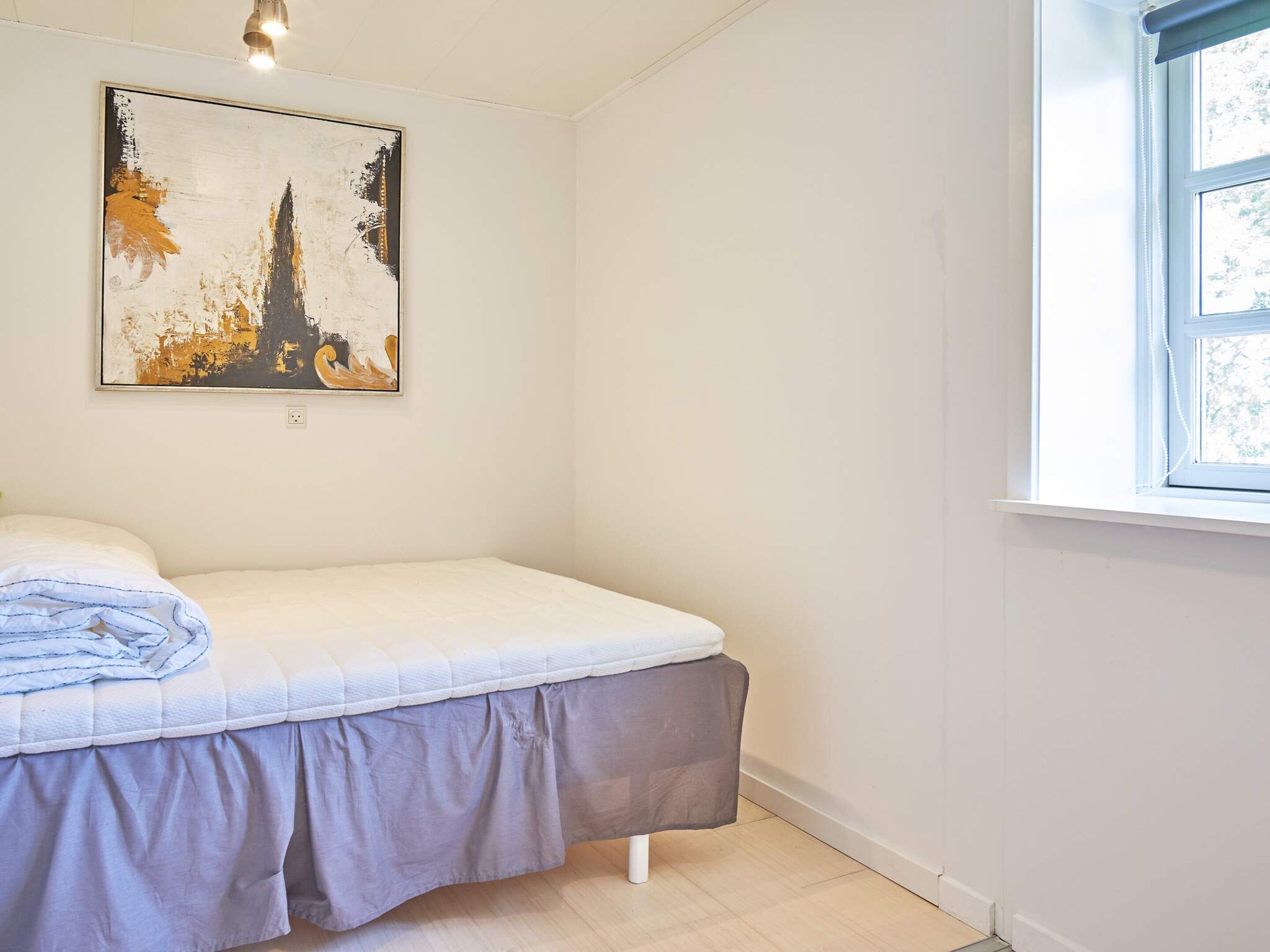 Uma cama de solteiro com saia cinza, colchão branco e cobertor azul. Há uma janela do lado direito e um quadro na parede.