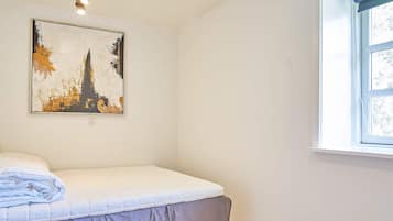 Uma cama de solteiro com saia cinza, colchão branco e cobertor azul. Há uma janela do lado direito e um quadro na parede.