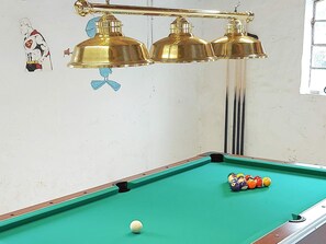 Uma mesa de bilhar com uma bola branca solitária e bolas coloridas, uma luminária de latão pendurada e uma parede com desenho do Superman.