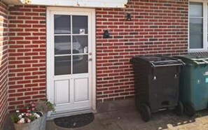 Uma casa de tijolos com porta branca e uma lixeira.