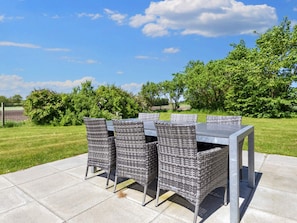 Pátio externo com mesa e cadeiras, cercado por vegetação e árvores.