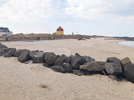 Uma praia arenosa com pedras grandes em primeiro plano, um pequeno edifício amarelo com telhado vermelho e algumas pessoas ao longe.