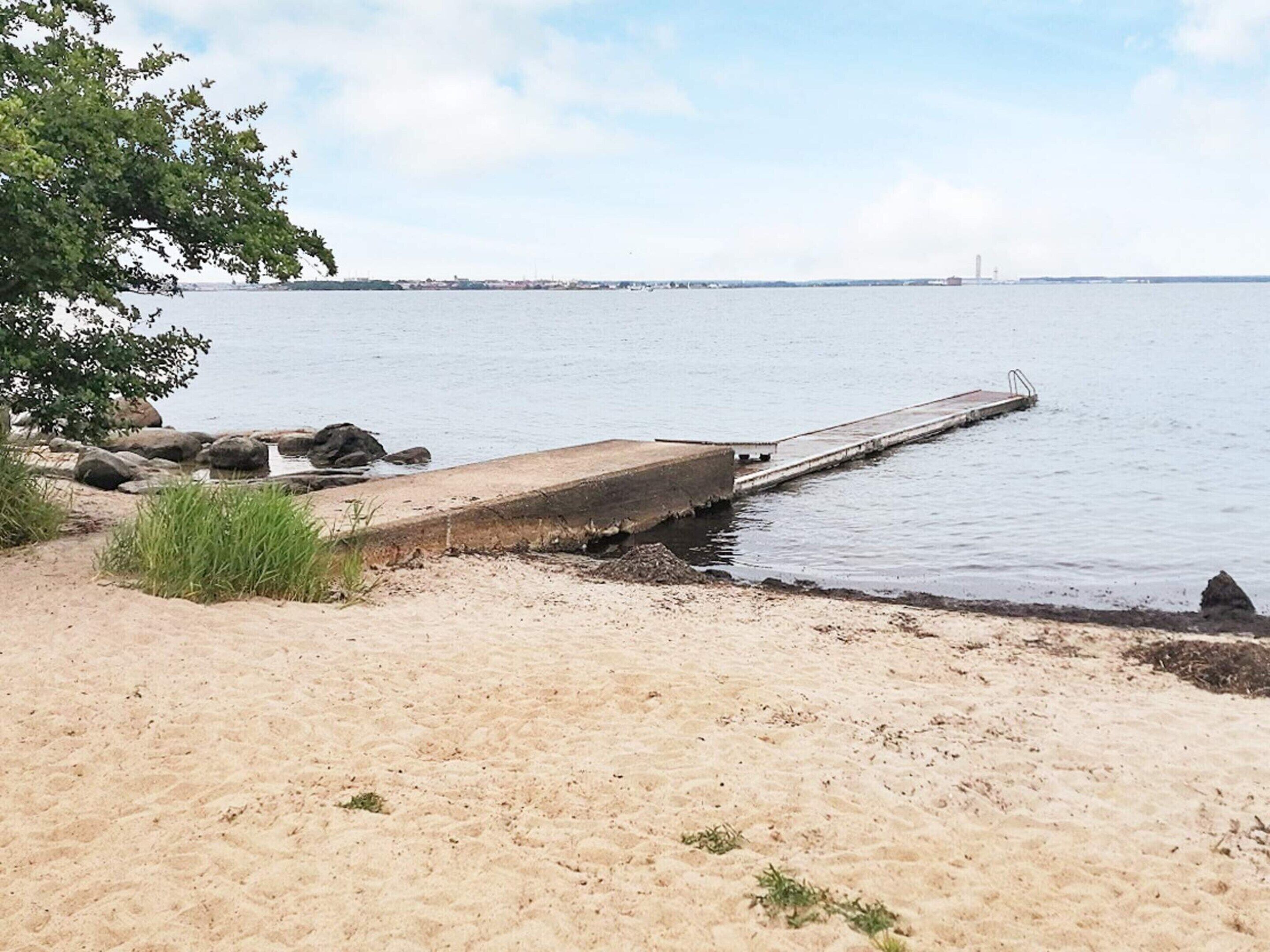 Uma praia arenosa com um cais de concreto que se estende por águas calmas, cercada por rochas e vegetação rala.