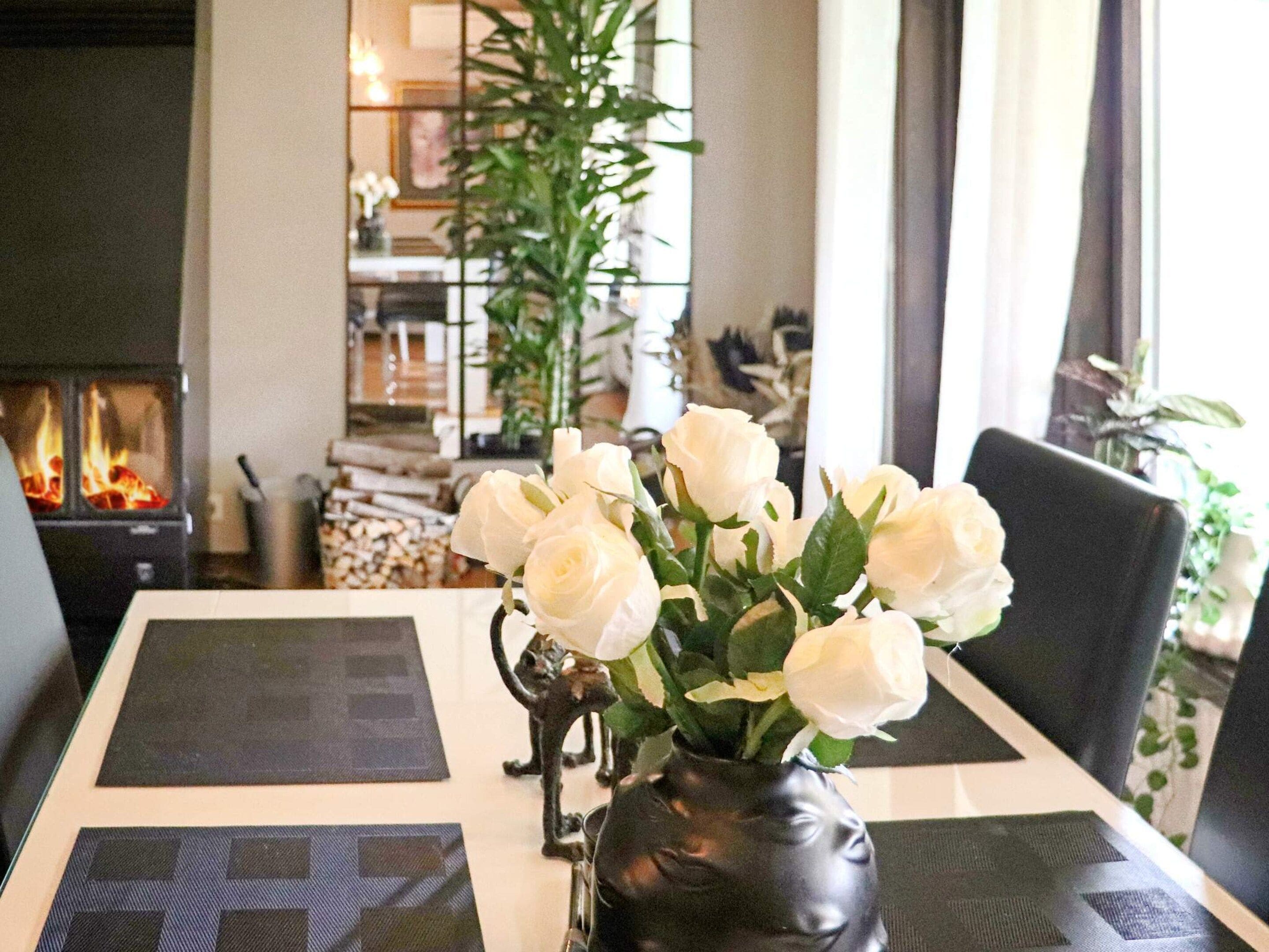 Uma mesa de jantar com um vaso de rosas brancas, um objeto decorativo preto e uma lareira ao fundo.
