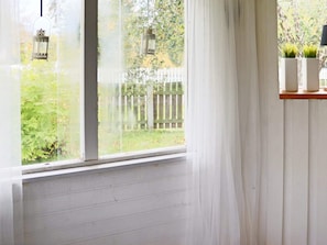 Quarto com uma janela grande, cortinas brancas, prateleira de madeira com plantas em vasos e parede branca.