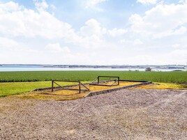 Uma área de cascalho com uma estrutura metálica, um campo verde e um corpo d'água ao longe.