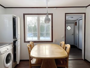 Cozinha equipada com máquina de lavar embutida, mesa de jantar com cadeiras, janela com vista para árvores e porta que dá para outro quarto.