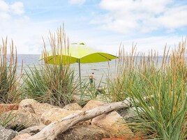 Uma praia com uma pessoa caminhando sob um guarda-sol verde, cercada por pedras e grama alta.