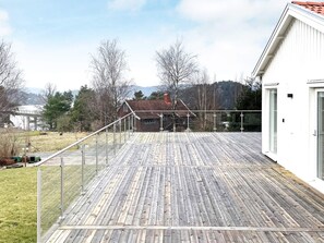 Um deck de madeira com guarda-corpo de metal, um edifício branco com portas de vidro e uma vista de uma casa com telhado vermelho ao fundo.