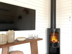Uma sala de estar com televisão de tela plana na parede, uma mesa de madeira com objetos decorativos e uma lareira a lenha preta com chamas visíveis.