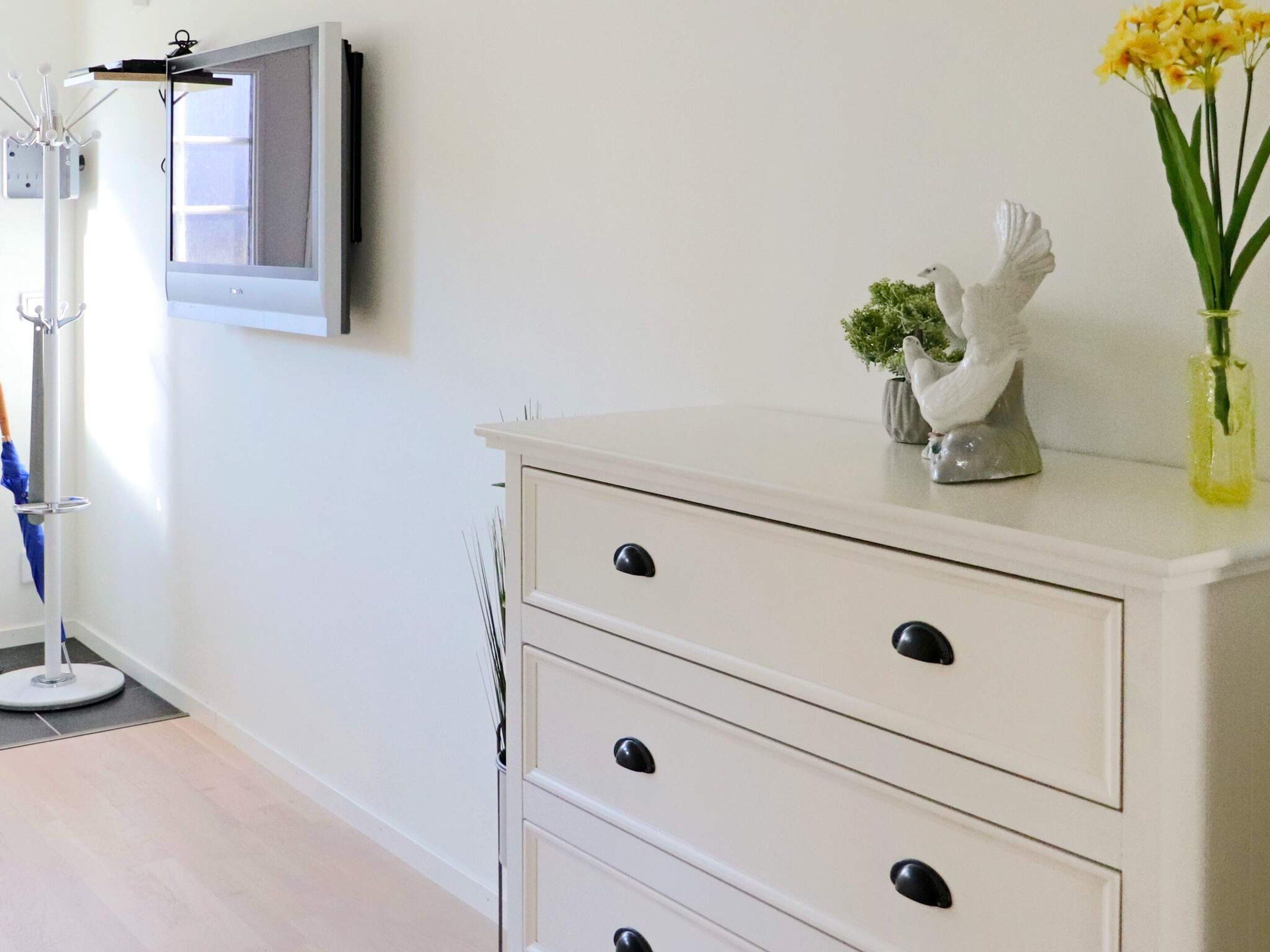 Quarto com um armário branco com gavetas, uma TV de tela plana na parede, um abajur e um vaso com flores sobre o armário.