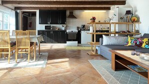 Cozinha moderna com armários pretos, fogão e micro-ondas. Há uma área de refeições com cadeiras de madeira e uma mesa. A sala de estar tem um sofá cinza e uma mesa de centro.