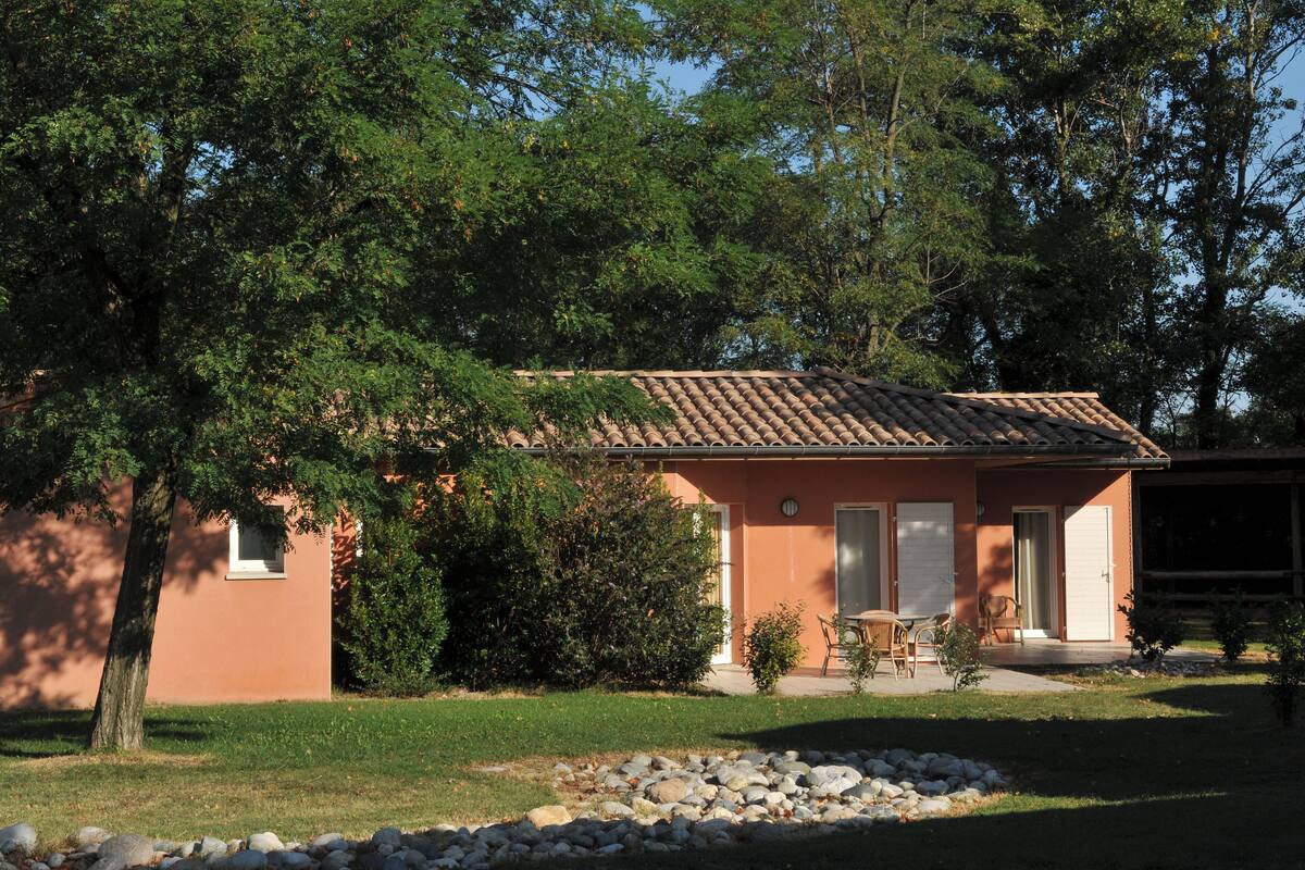 Uma casa térrea com telhado de telhas, um pequeno alpendre com mesa e cadeiras, e um jardim com um canteiro de pedras.