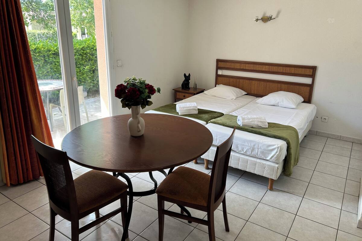 Quarto de hotel com uma cama, uma mesa com cadeiras, um vaso com flores e vista para o verde lá fora.