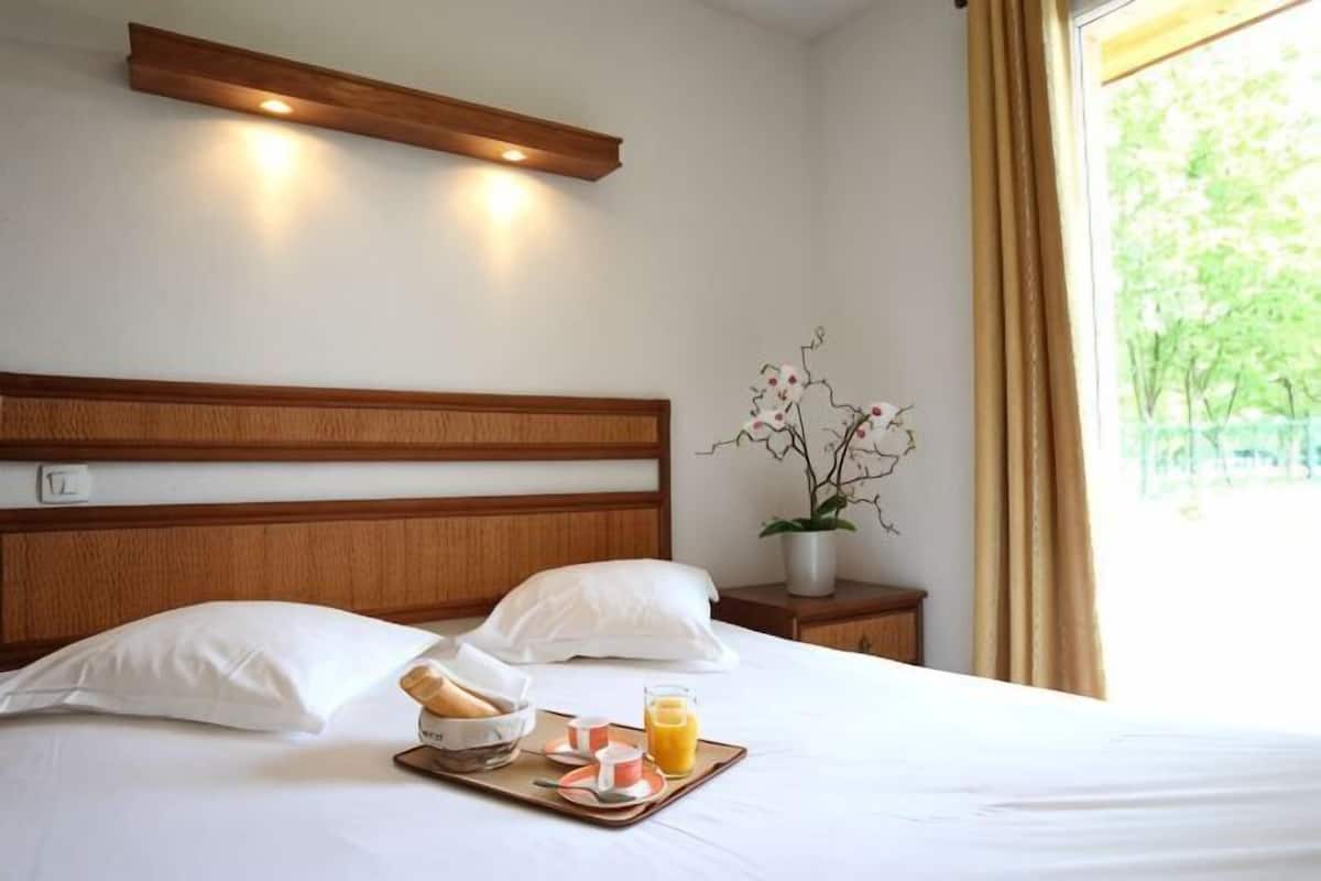 Quarto de hotel com cama, almofadas, bandeja de café da manhã, mesa de cabeceira, planta em vaso e janela com cortinas.