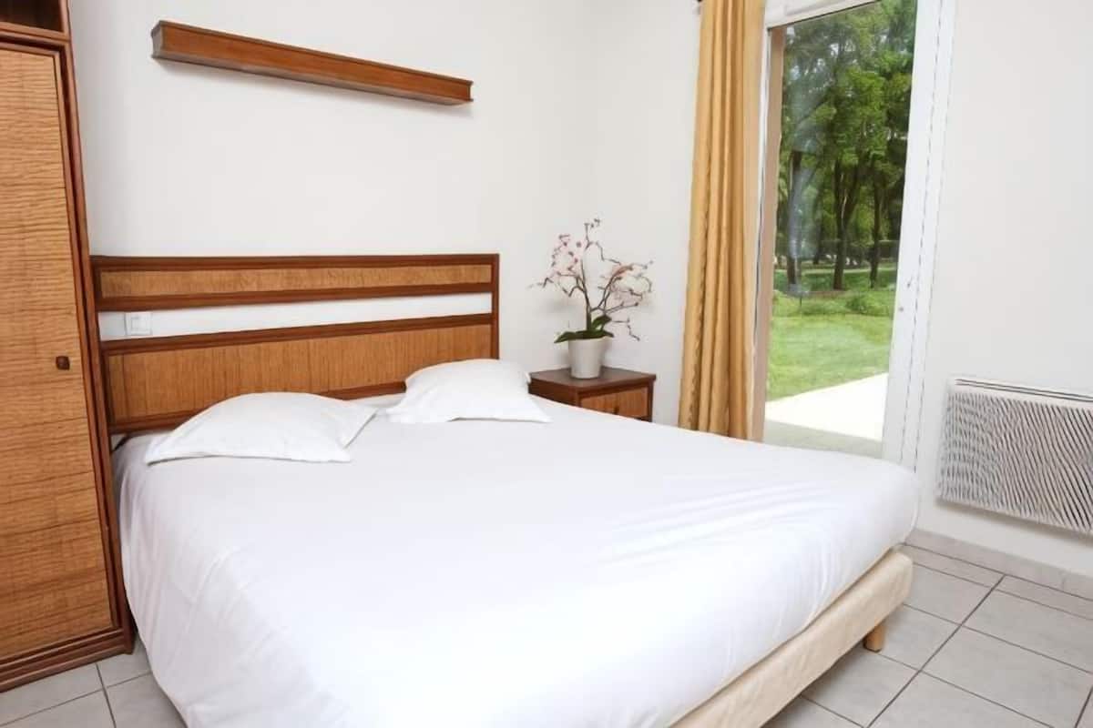 Quarto com cabeceira de madeira, cama com roupa de cama branca, mesa de cabeceira com planta em vaso e vista para o verde lá fora.