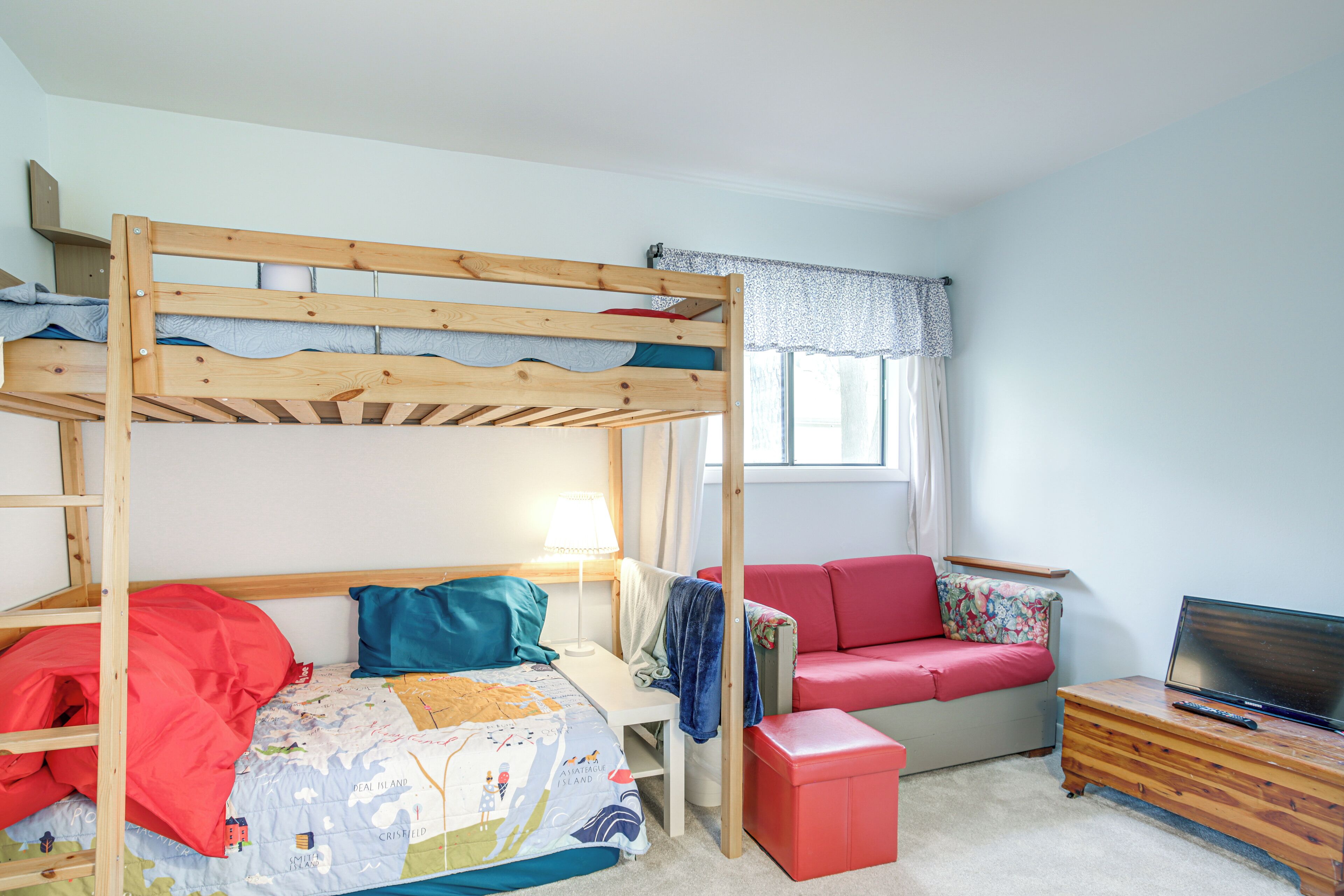 Uma cama beliche com uma escrivaninha e um sofá vermelho em um quarto com janela e uma televisão sobre um aparador de madeira.