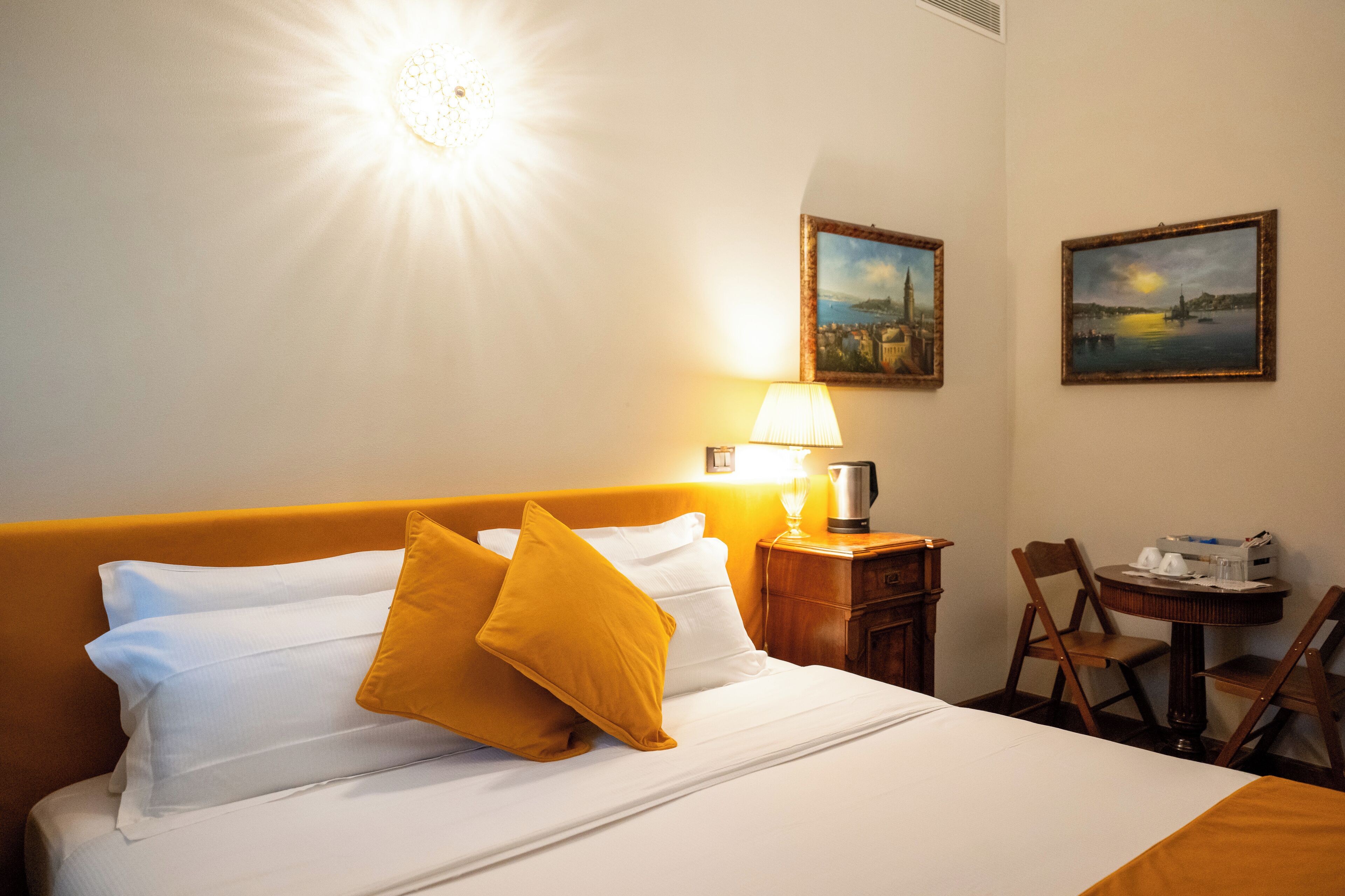 Quarto de hotel com uma cama, uma mesa de cabeceira com abajur, duas cadeiras e duas pinturas na parede.