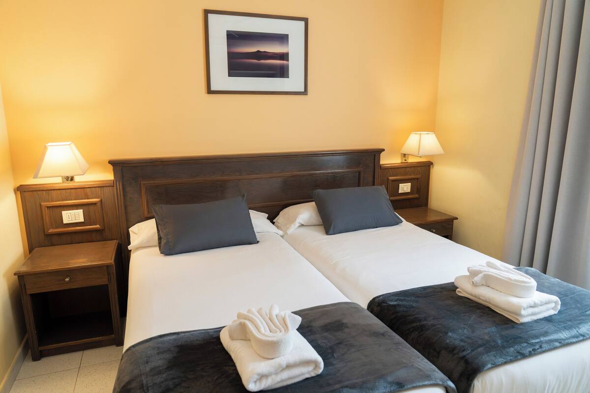 Quarto de hotel com duas camas, mesas de cabeceira com luminárias, um quadro na parede e toalhas nas camas.