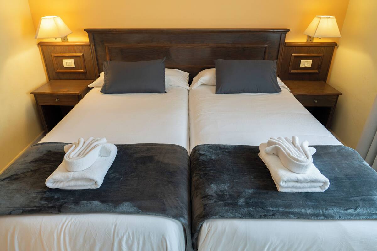 Uma cama de casal com duas almofadas, uma mesa de cabeceira com abajur e uma toalha em cada cama.