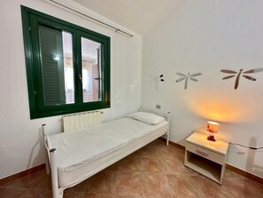 Quarto pequeno com uma cama de solteiro, janela, mesa de cabeceira com luminária e detalhes decorativos nas paredes.
